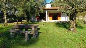 Uma casa rústica com telhado de telhas, rodeada por árvores e um espaço gramado com um banco de pedra.