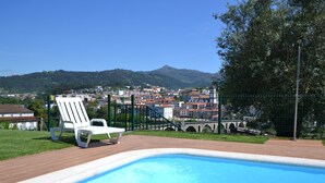 Uma piscina com uma espreguiçadeira branca, uma cerca verde e vista para uma cidade e montanhas ao fundo.
