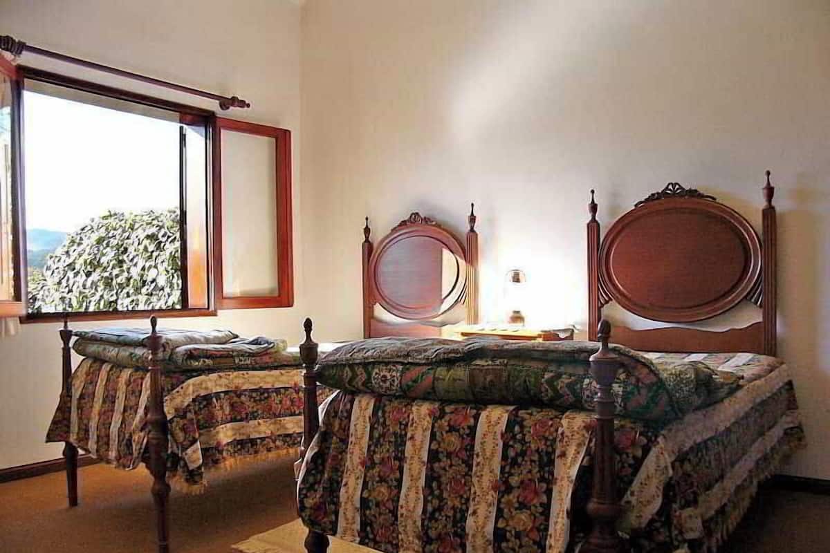 Quarto com duas camas de madeira com roupa de cama floral, uma janela com vista e uma mesa de cabeceira com abajur.