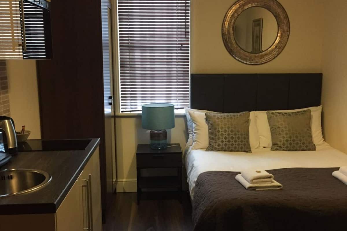 Quarto de hotel com uma cama, uma mesa de cabeceira com luminária, um espelho e uma área de cozinha com pia.
