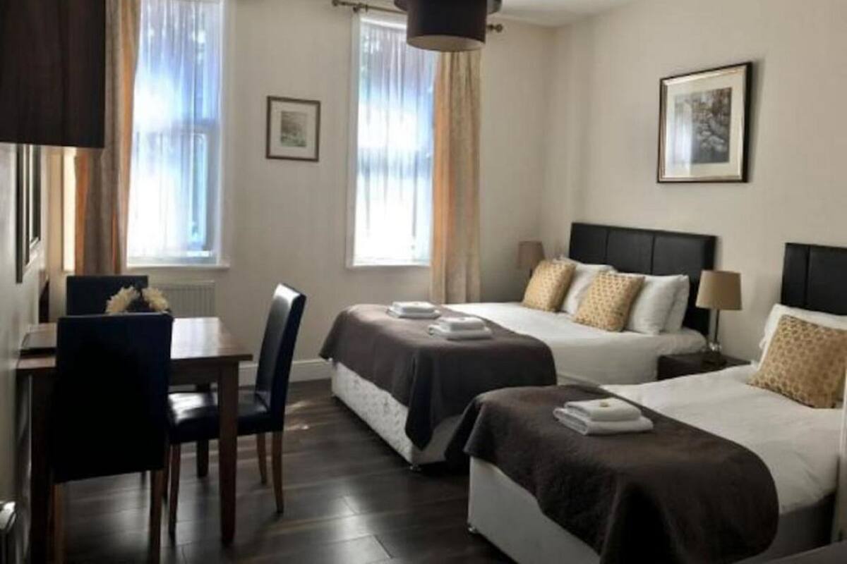 Quarto de hotel com duas camas, uma escrivaninha e uma cadeira. Há cortinas nas janelas e um quadro na parede.