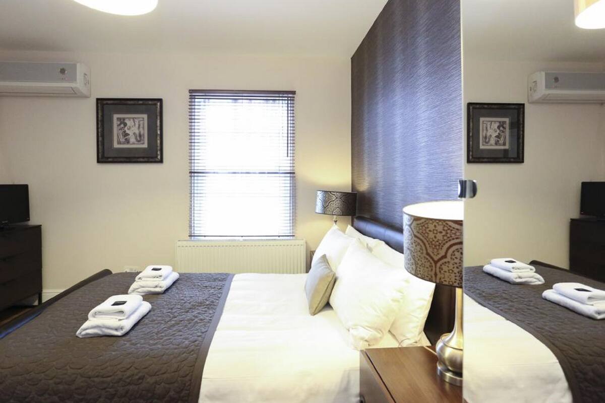 Quarto de hotel com cama grande, duas mesas de cabeceira, abajur e janela com persianas.