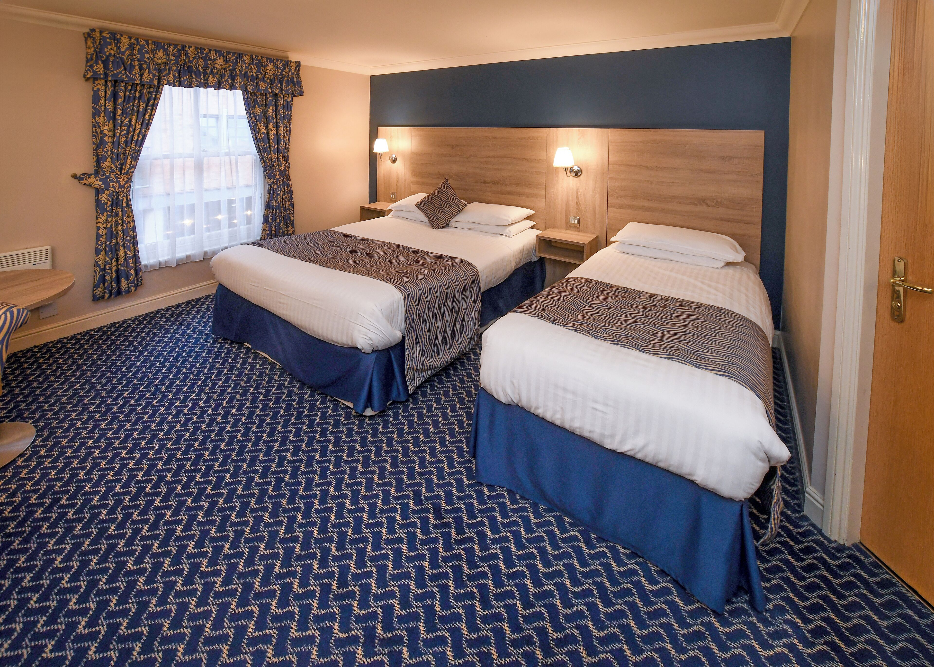 Quarto de hotel com duas camas, uma cabeceira de madeira, uma mesa de cabeceira com abajur e um tapete estampado.