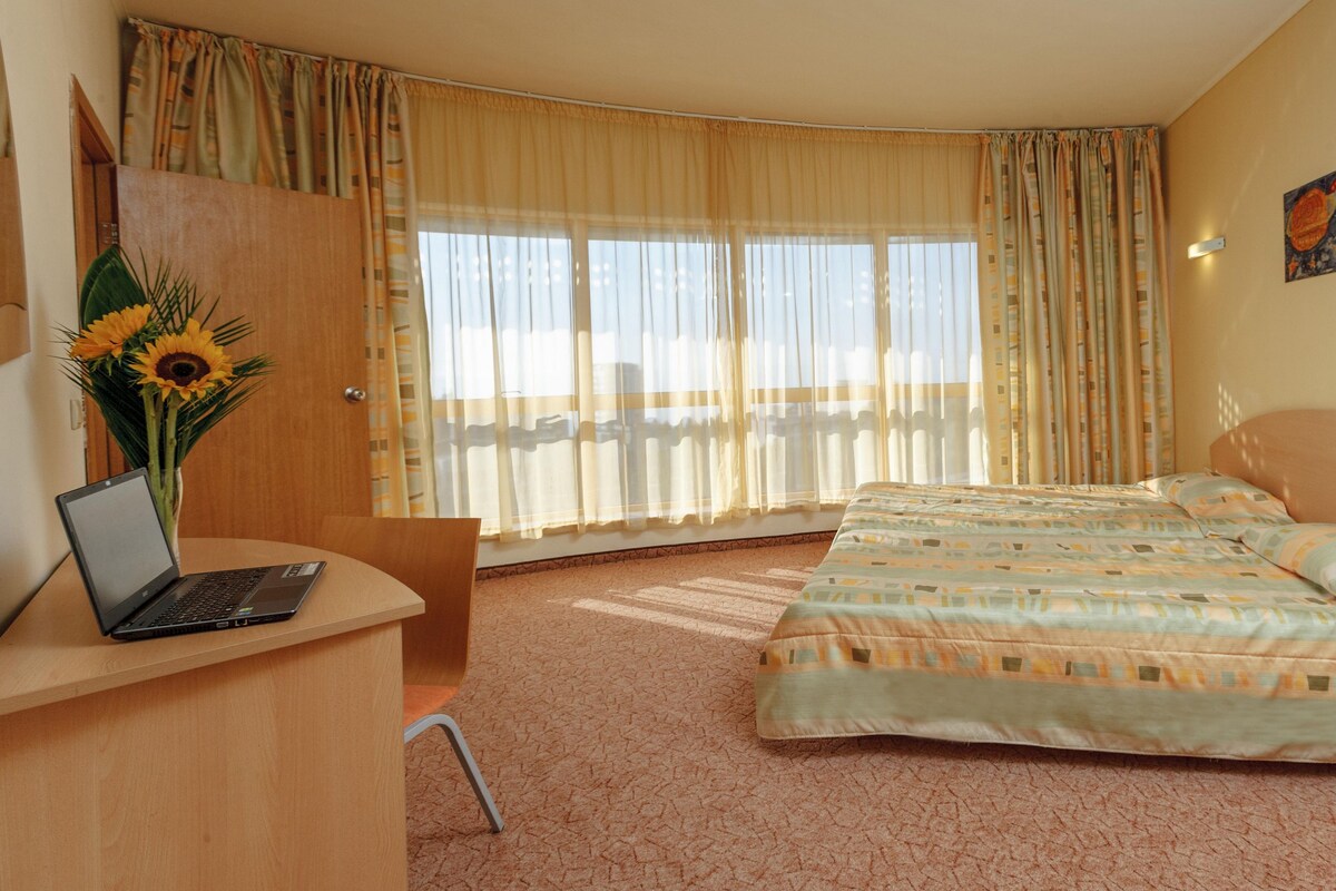 Quarto de hotel com uma cama, uma escrivaninha com um laptop, uma cadeira, um telefone e um quadro na parede.