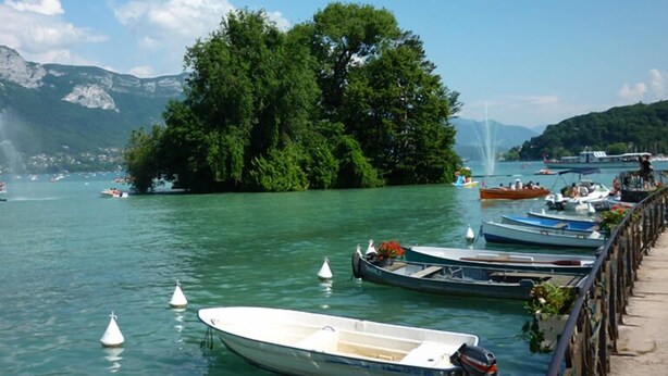 Um lago com barcos atracados na margem, uma fonte na água e uma ilha arborizada ao fundo.