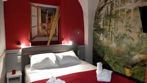 Quarto de hotel com parede vermelha, cama com roupa de cama branca, mesa de cabeceira com vela vermelha e um mural de paisagem tropical.