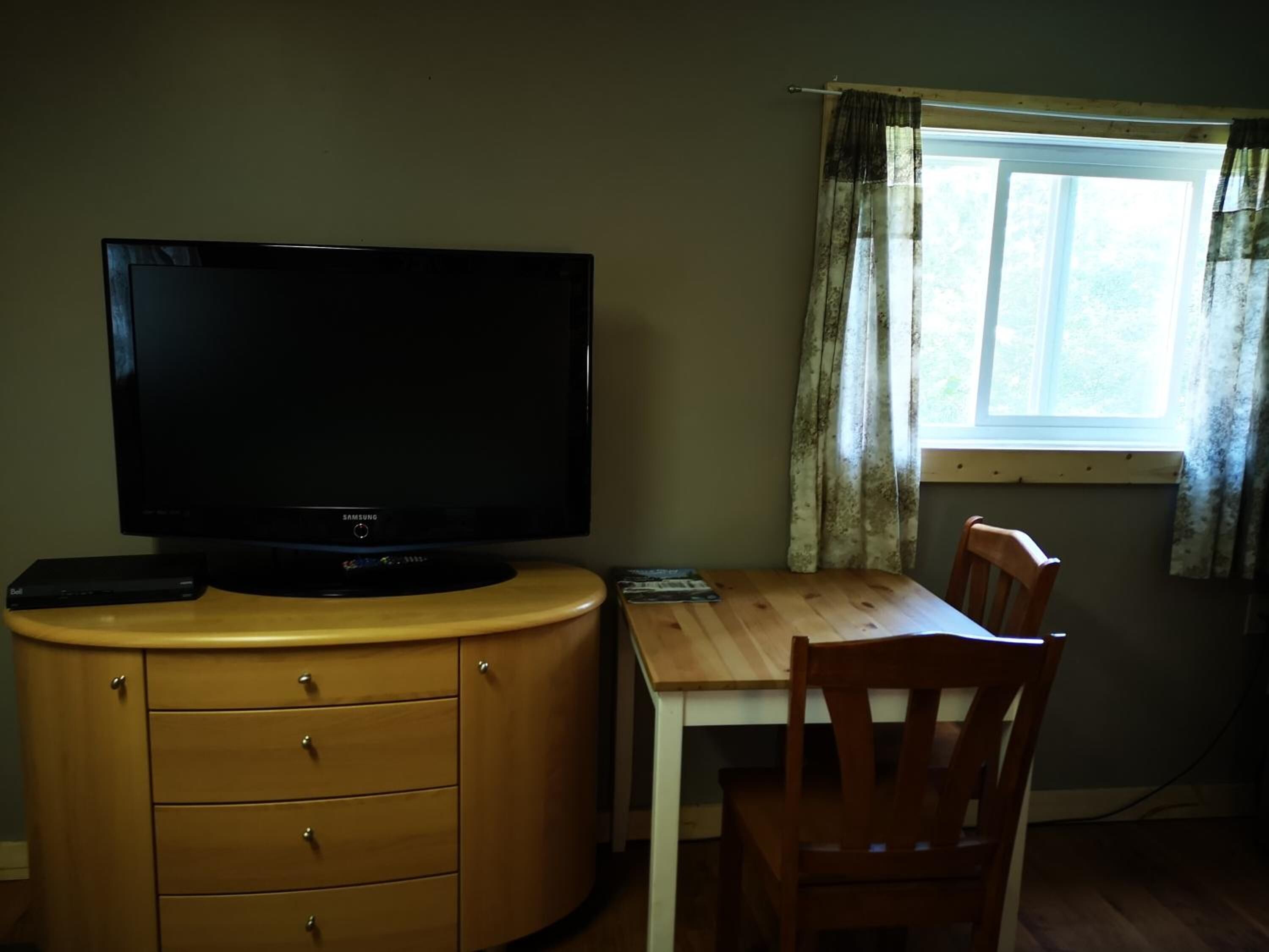 Quarto com televisão sobre um armário de madeira, uma mesa de madeira e uma cadeira.