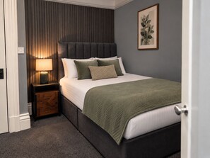 Quarto com cama, mesa de cabeceira, abajur e um quadro na parede.