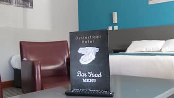 Quarto de hotel com um expositor de menu de bar, uma cama, uma cadeira vermelha e uma parede com uma fotografia a preto e branco.