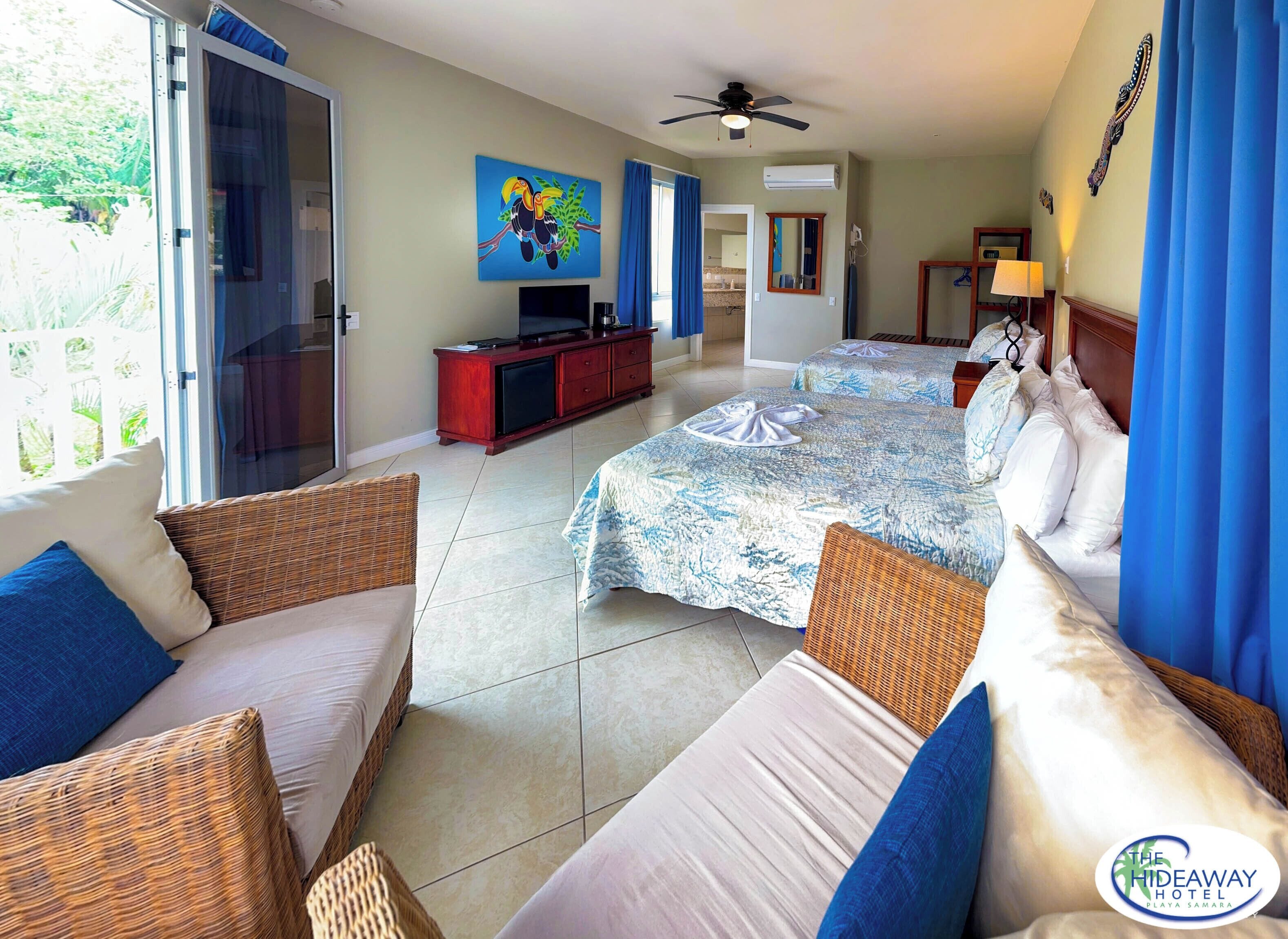 Quarto de hotel com duas camas, móveis de vime, ventoinha no teto e uma pintura de tucano.