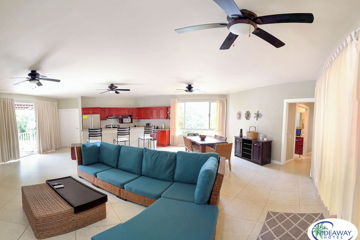 Uma sala de estar ampla com um sofá azul, uma mesa de centro e uma mesa de jantar. Há ventiladores de teto, uma cozinha com armários vermelhos e vista para o exterior.