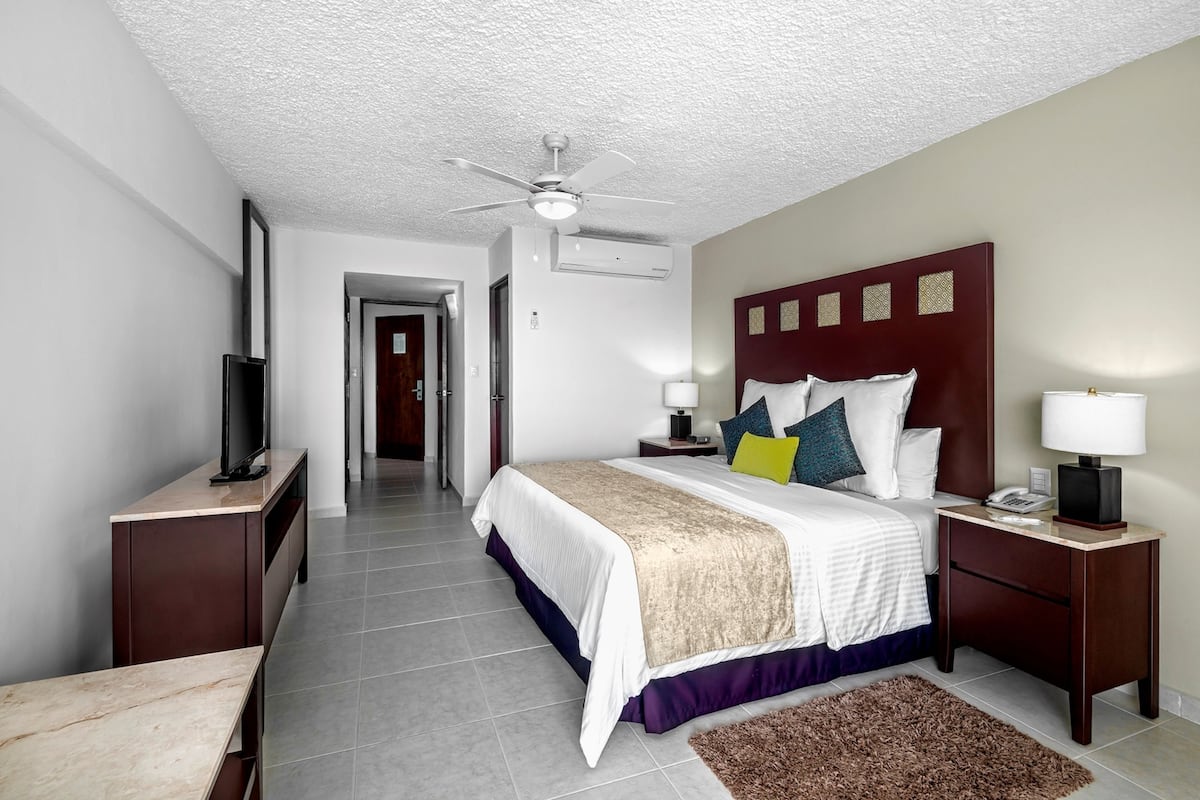Quarto de hotel com cama grande, ventoinha no teto, televisão em um suporte e mesa de cabeceira com luminária.