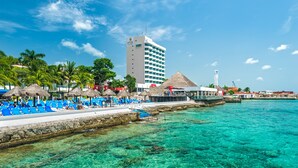 Um hotel à beira-mar com águas turquesa cristalinas, área de piscina com cadeiras azuis e um edifício alto ao fundo.
