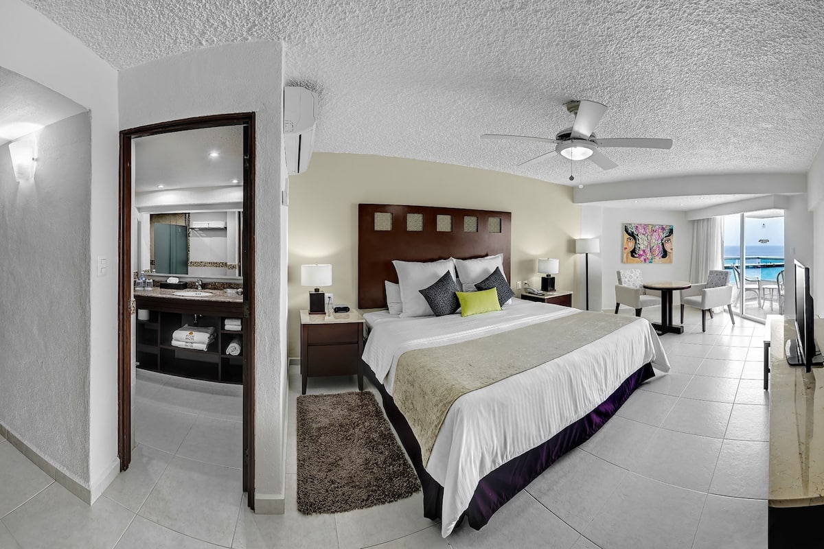 Quarto de hotel moderno com uma cama grande, um ventilador de teto, vista para o oceano e banheiro com toucador e espelho.