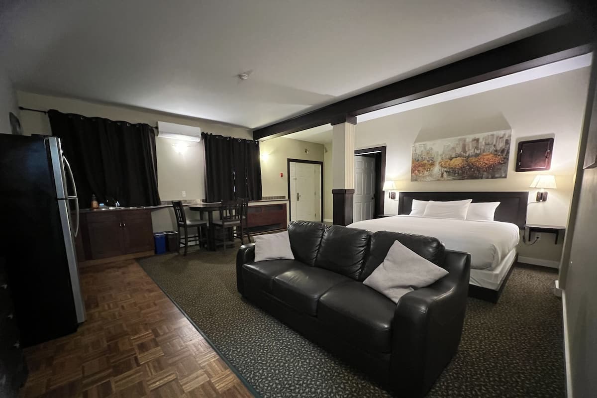 Quarto de hotel com uma cama, um sofá de couro preto, uma sala de jantar com mesa e cadeiras, e uma cozinha com geladeira.