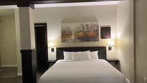 Quarto de hotel com uma cama grande, duas luminárias de cabeceira, um quadro com vista para a cidade na parede e um espaldar escuro.