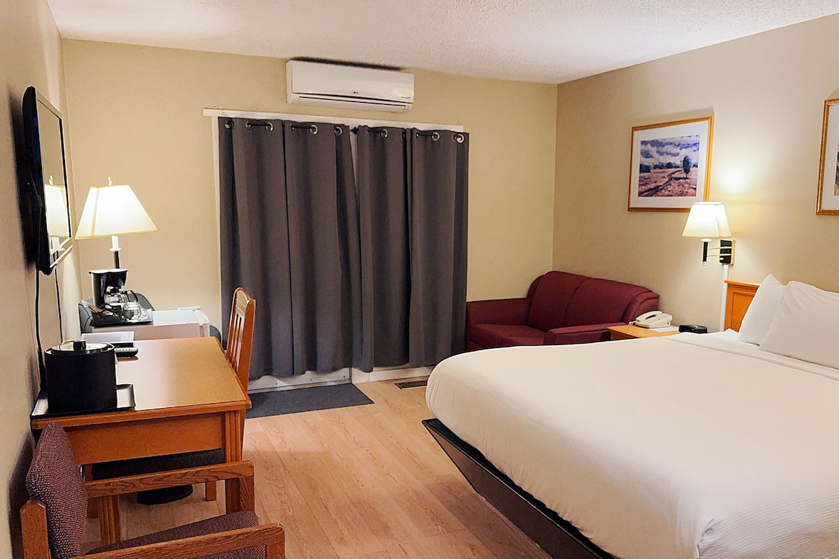 Quarto de hotel com cama, escrivaninha com luminária, cadeira, sofá vermelho, telefone e duas pinturas na parede.