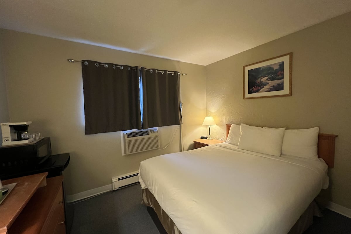 Quarto de hotel com cama, escrivaninha com cafeteira, luminária, quadro na parede e janela com cortinas.