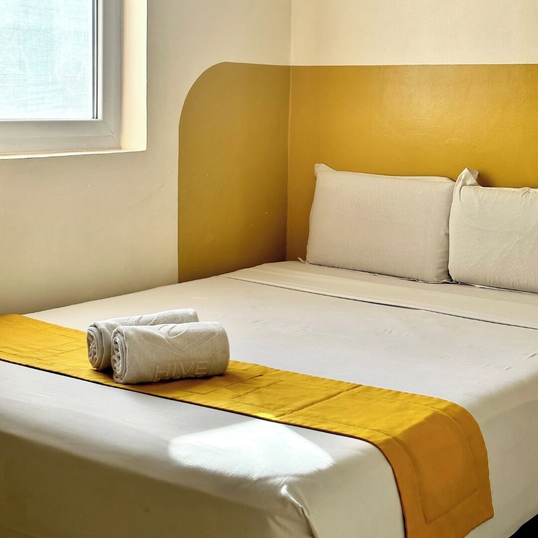 Quarto de hotel com cama, janela e uma parede de destaque amarela.