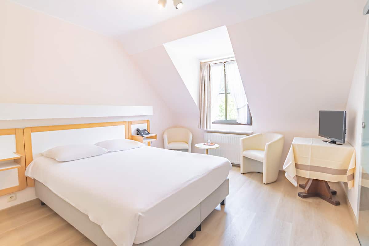 Quarto de hotel moderno com uma cama grande, uma escrivaninha com televisão, duas cadeiras e uma mesinha.