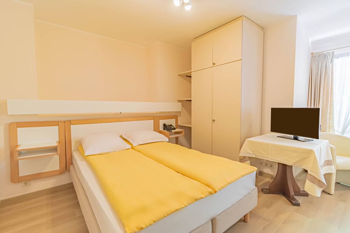 Quarto de hotel com uma cama de casal, um guarda-roupa, mesa de cabeceira com abajur e televisão.