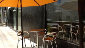 Um café com mesas e cadeiras ao ar livre, e um grande guarda-sol laranja oferecendo sombra.