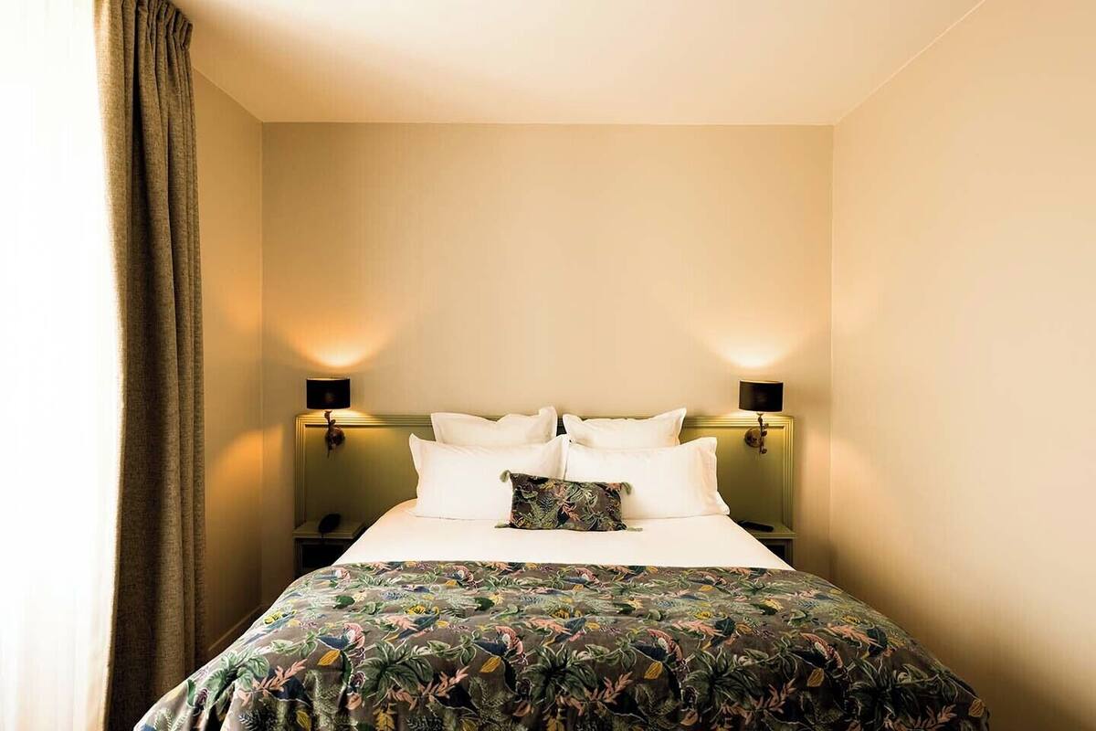 Quarto de hotel com cama, duas luminárias de cabeceira, colcha florida e cortina.