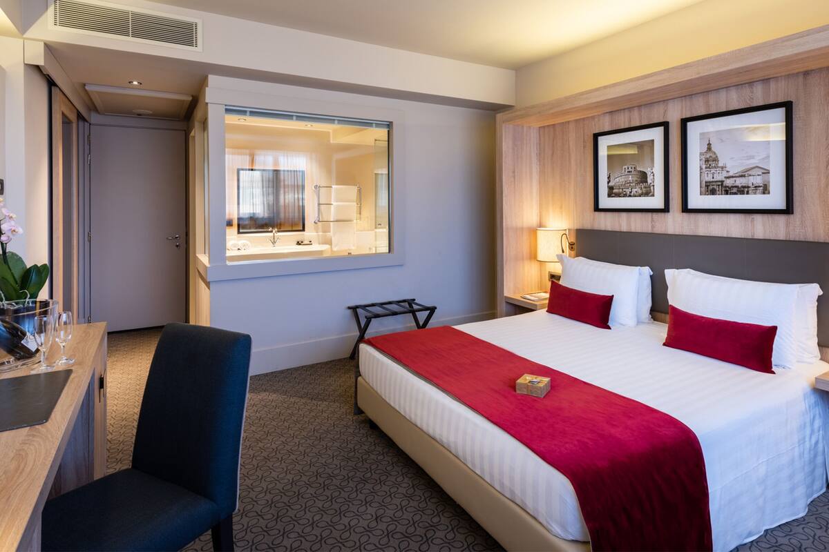 Quarto de hotel com uma cama grande, duas mesas de cabeceira, uma escrivaninha com cadeira, um espelho e duas molduras na parede.