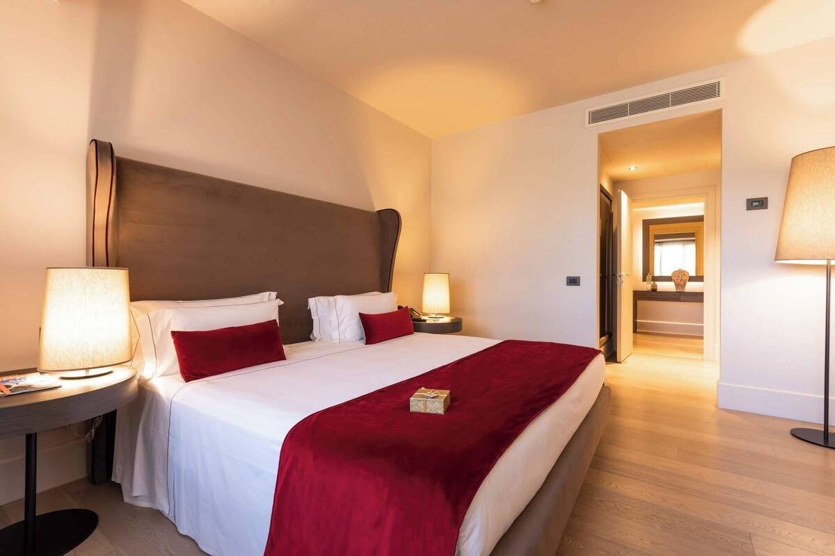 Quarto de hotel com uma cama grande, duas mesas de cabeceira com luminárias, um cobertor vermelho e uma cabeceira de madeira.