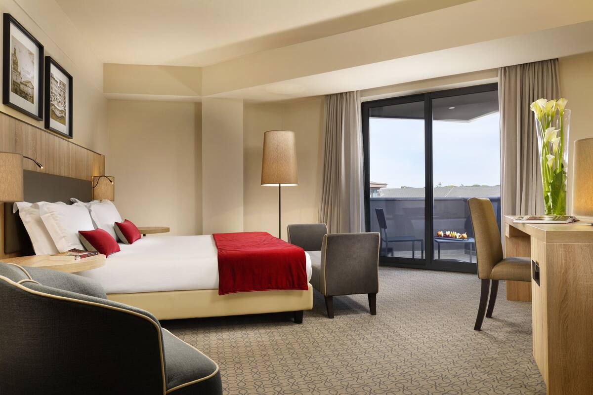 Quarto de hotel com cama, escrivaninha, cadeira, abajur e uma janela grande com vista.