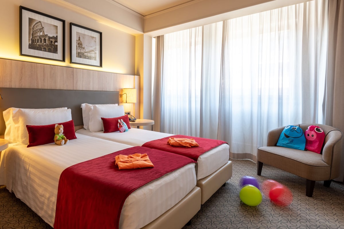 Quarto de hotel com duas camas, uma mesa redonda, uma cadeira e uma janela grande com cortinas.