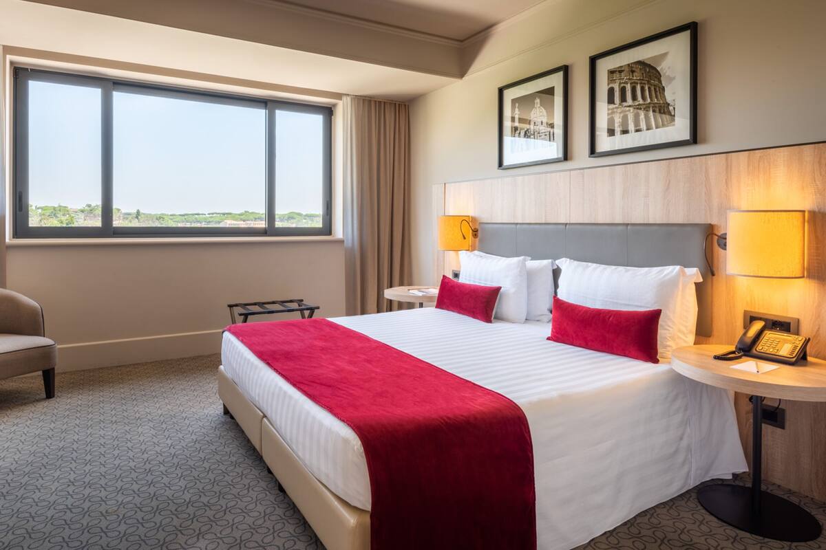 Quarto de hotel com uma cama grande, duas luminárias de cabeceira, uma cadeira, janela com vista e duas imagens emolduradas na parede.
