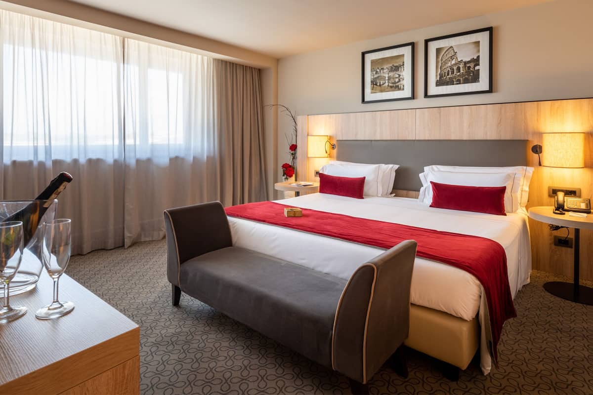 Quarto de hotel com uma cama, mesas de cabeceira, escrivaninha com cadeira, uma janela grande com cortinas e duas imagens emolduradas na parede.