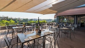 Uma área de refeições ao ar livre com mesas e cadeiras, um toldo grande e vista para a paisagem urbana.
