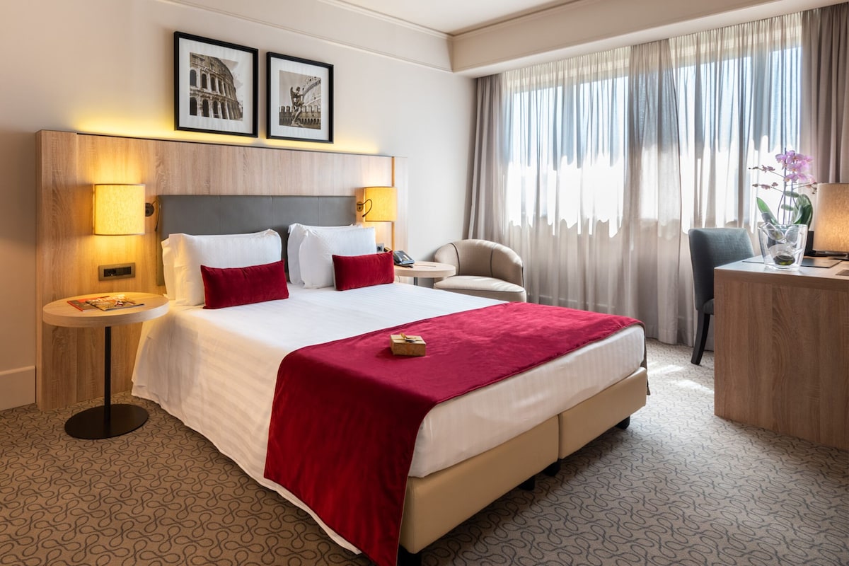 Quarto de hotel com uma cama grande, mesa de cabeceira, cadeira, escrivaninha e janela com cortinas.