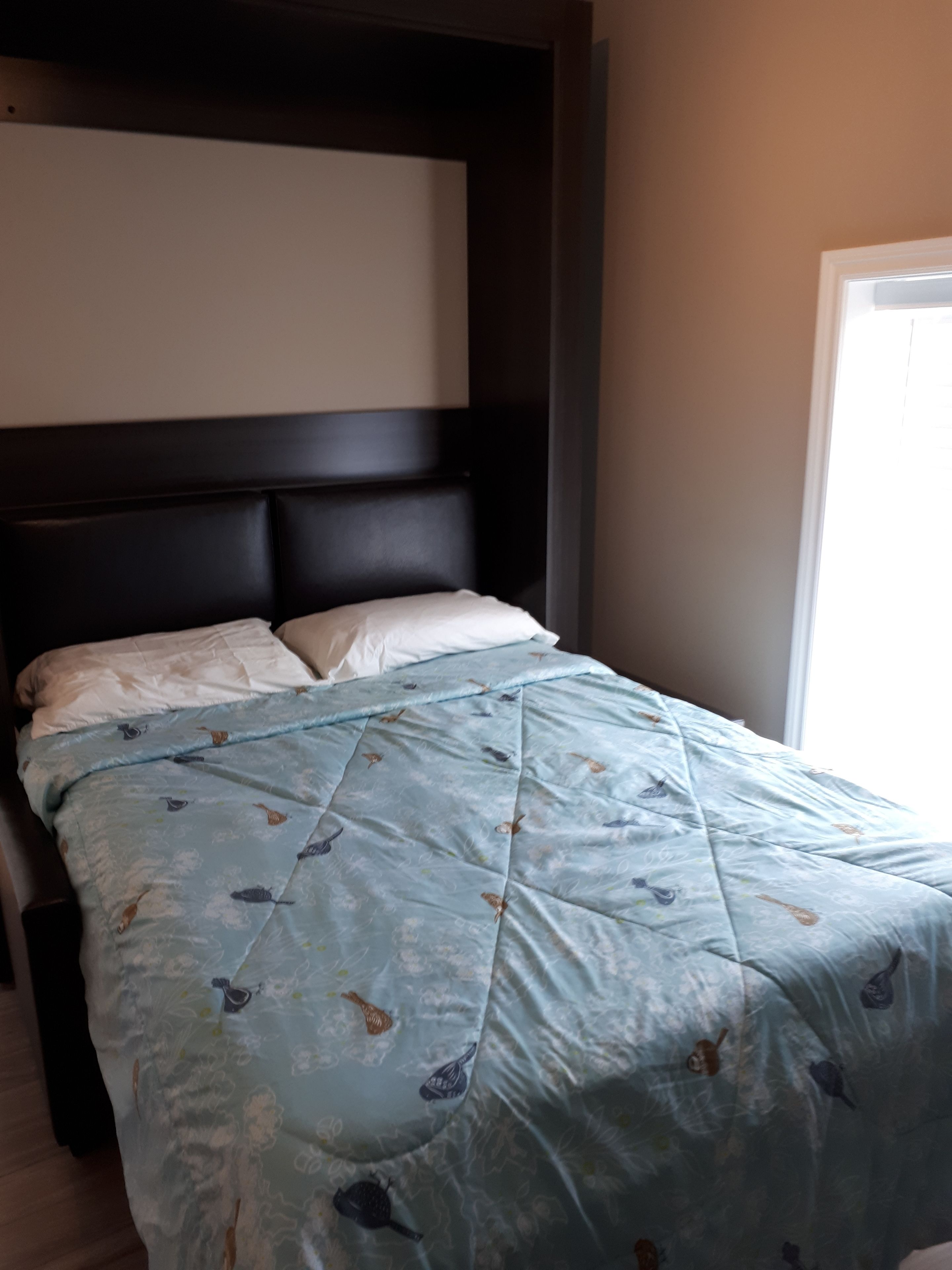 Uma cama com cabeceira escura, um edredom azul claro com estampas de pássaros e um travesseiro branco.