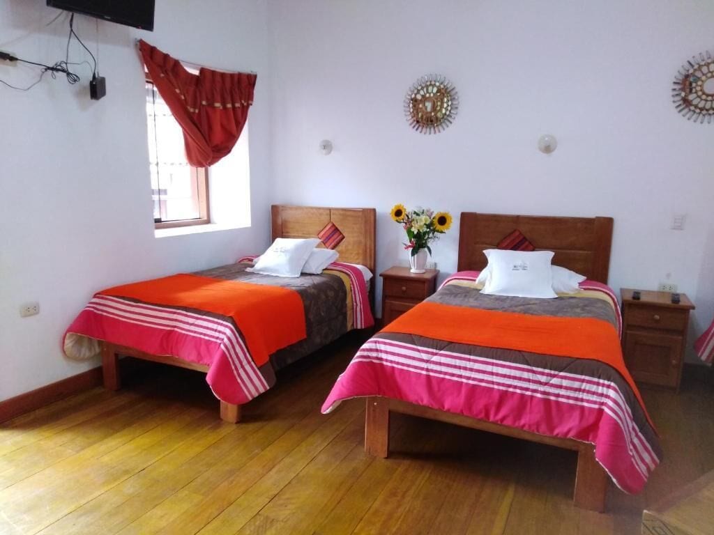 Quarto com duas camas de madeira, cada uma com colcha e almofadas listradas em vermelho e branco. Há uma pequena mesa de cabeceira com um vaso de flores entre as camas.