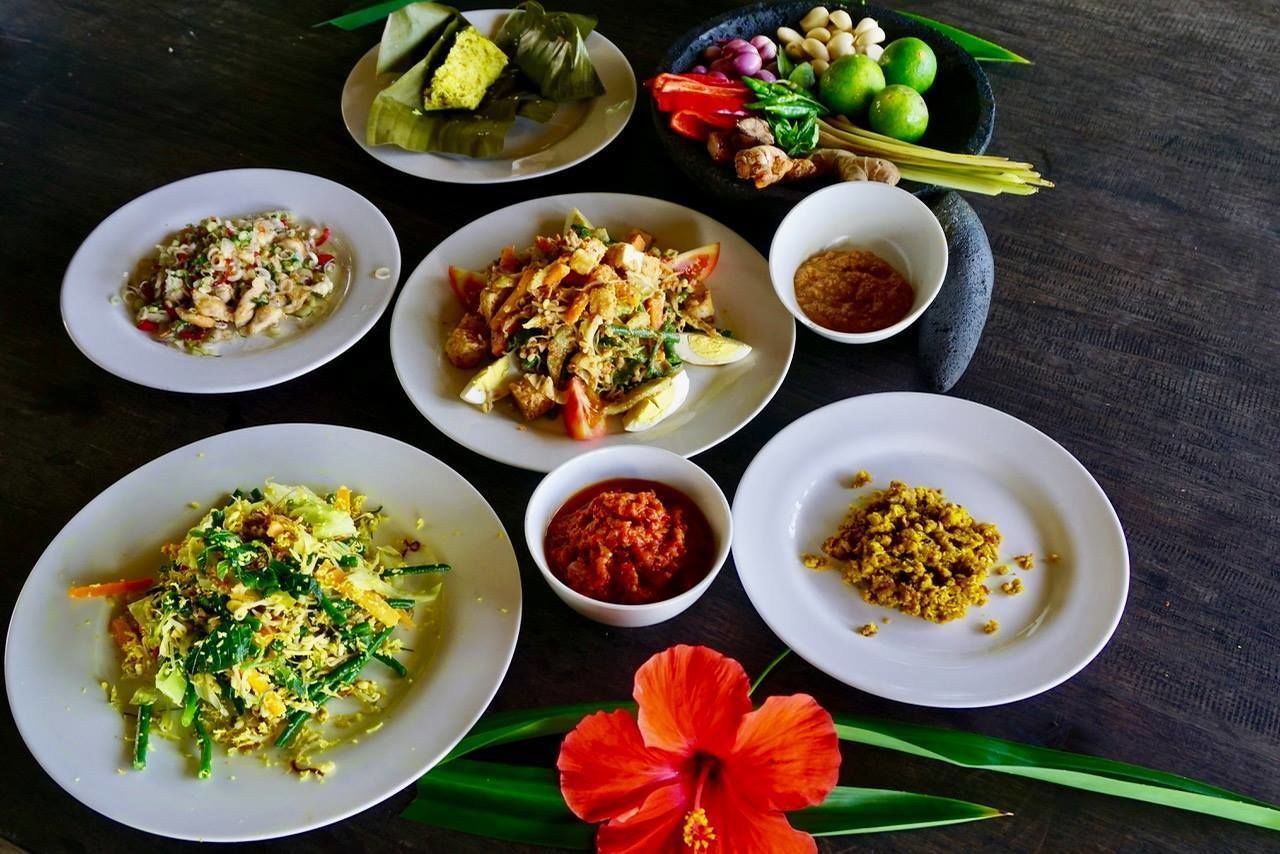 Uma mesa com diversos pratos, incluindo uma salada, um prato com carne e legumes, e um prato com molho vermelho. Há também algumas frutas e uma flor sobre a mesa.