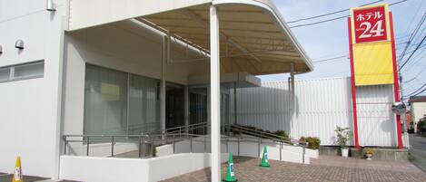 Um edifício moderno com entrada coberta, grandes janelas e uma placa vermelha e amarela bem visível que indica serviço 24 horas.