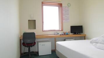 Quarto de hotel pequeno com uma cama de solteiro, uma escrivaninha com computador, uma cadeira, um refrigerador pequeno, uma janela e um calendário na parede.