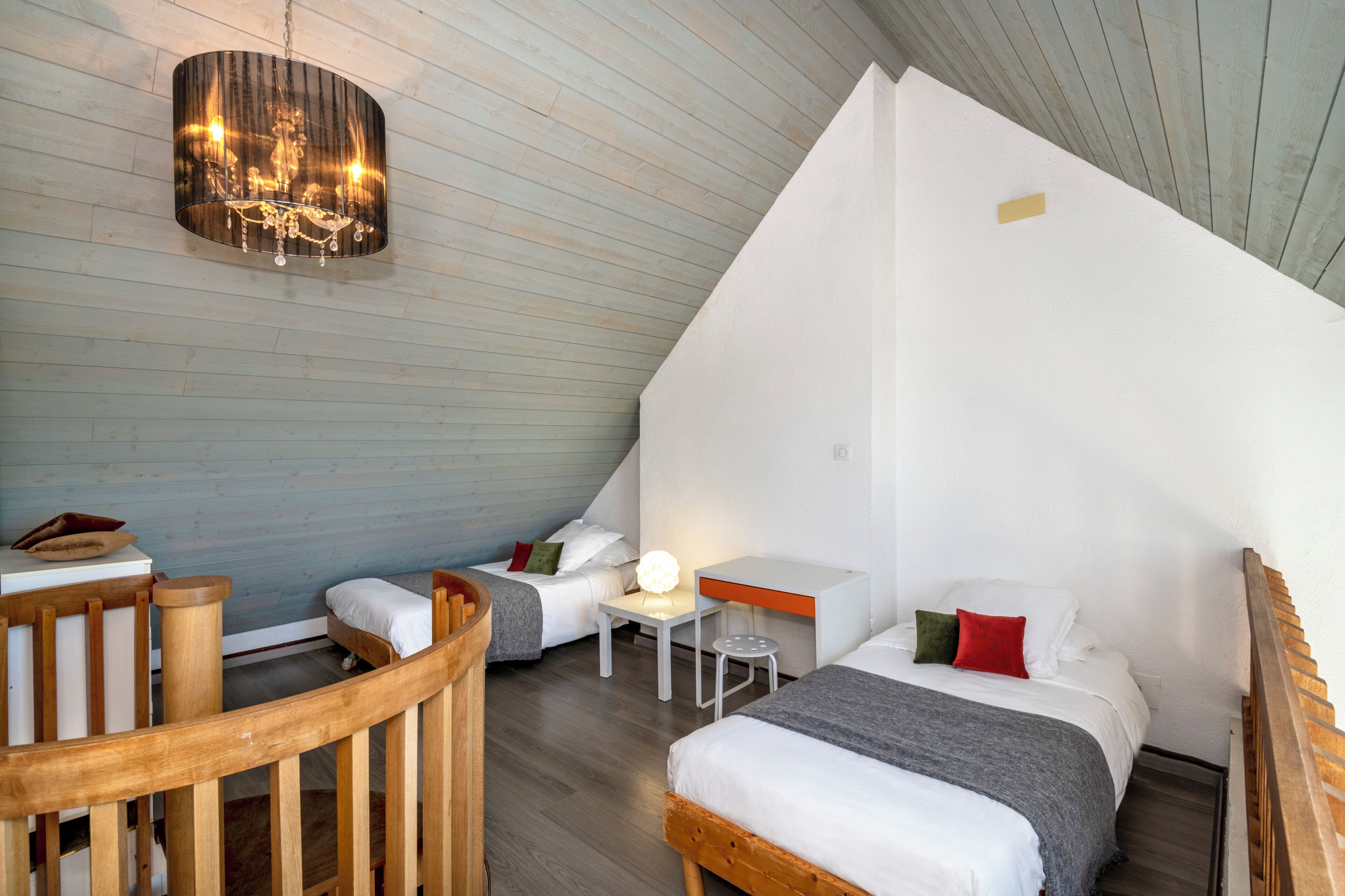 Quarto moderno e bem iluminado com teto inclinado, uma cadeira de madeira, uma mesa pequena e duas camas com almofadas.