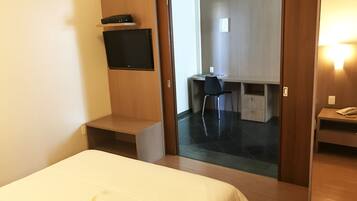 Quarto de hotel com cama, escrivaninha, cadeira, televisão e telefone em mesinha de cabeceira.