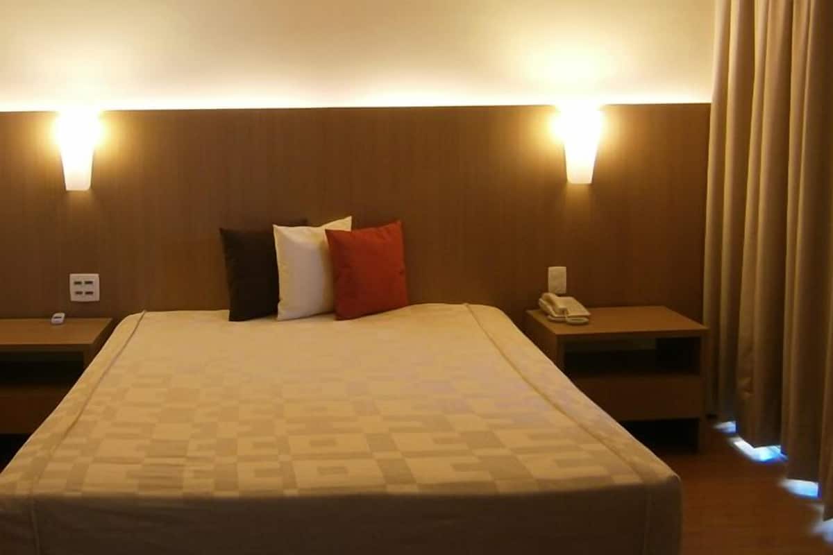 Quarto de hotel com uma cama grande, duas mesas de cabeceira e uma luminária fixada na parede.