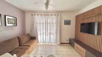 Sala de estar com sofá, televisão fixada em parede de madeira, ventilador de teto e janela com cortinas.