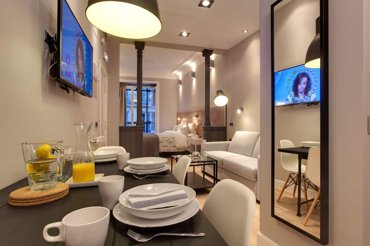 Quarto de hotel moderno com área de refeições, cama e TV exibindo uma pessoa.
