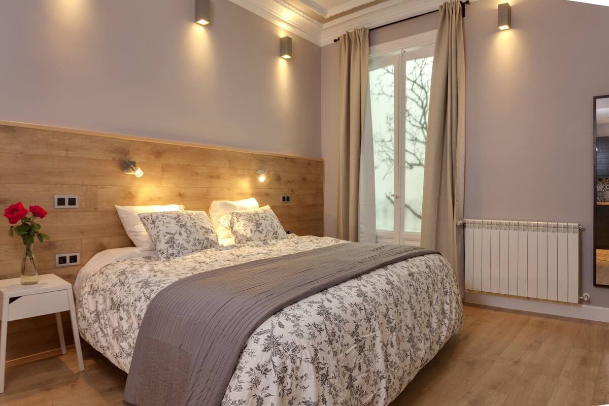 Quarto com cabeceira de madeira, cama grande com roupa de cama florida, abajures, mesa de cabeceira com um vaso de flores e uma janela com cortinas.