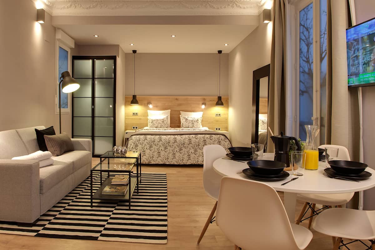Quarto de hotel moderno com sofá, mesa de jantar com cadeiras, cama com cabeceira, televisão e janela com cortinas.