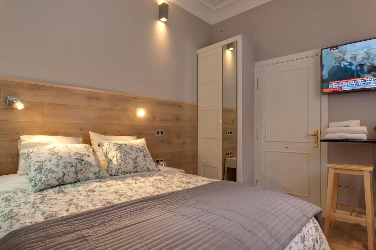 Quarto com cabeceira de madeira, cama com roupa de cama floral, TV fixada na parede e porta de acesso a um banheiro.
