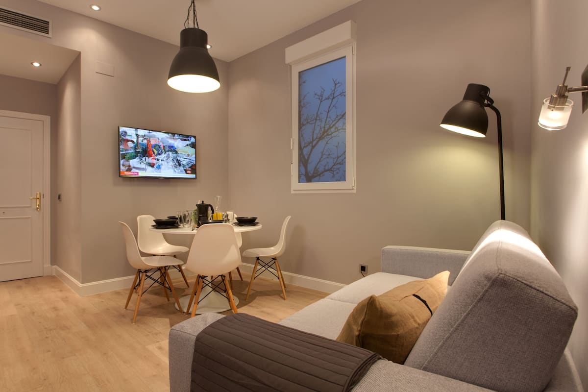 Uma sala de estar moderna com um sofá cinzento, uma mesa de jantar com cadeiras brancas, uma televisão de tela plana na parede e uma janela com vista para uma árvore.