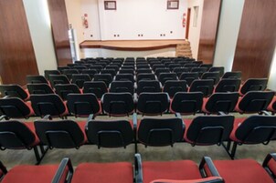 Uma sala de conferências com fileiras de cadeiras pretas com almofadas vermelhas, um palco na frente e um extintor de incêndio na parede.